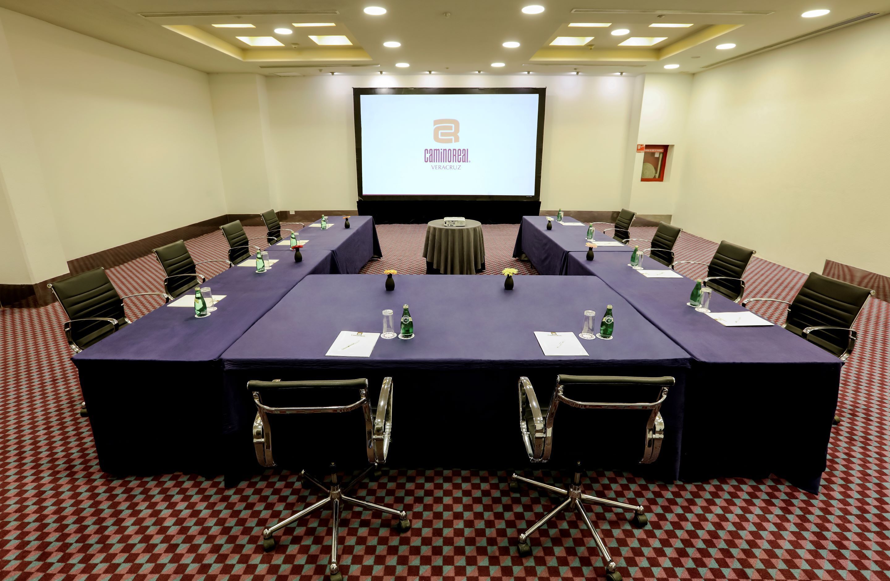 Sala de conferência com uma tela grande exibindo um logotipo, várias mesas com cadeiras e piso coberto de carpete.