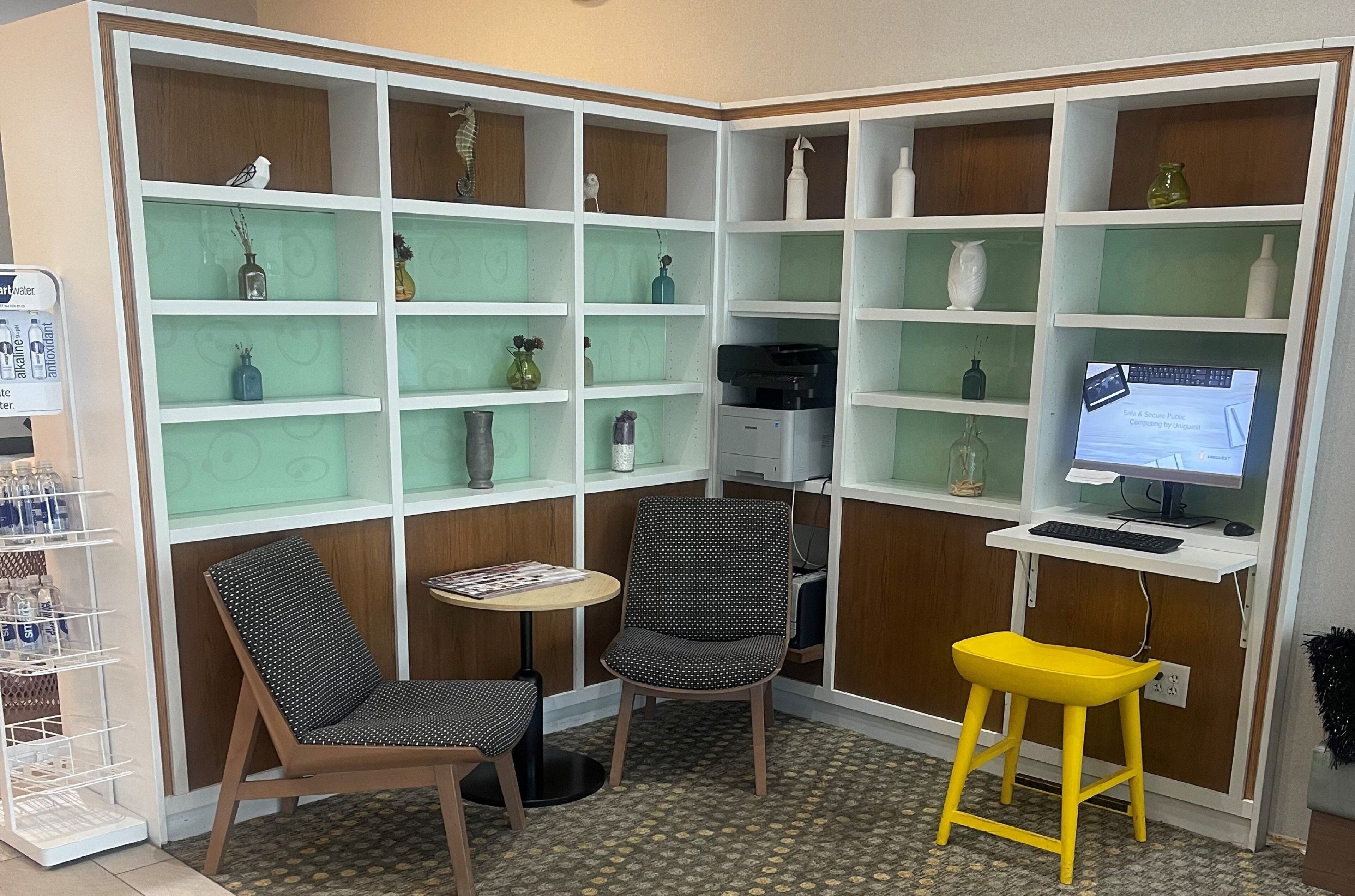 Quarto de canto com duas cadeiras, uma mesa pequena, um banquinho amarelo e um computador em uma prateleira. Prateleiras repletas de objetos decorativos.