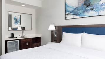 Quarto de hotel com cama, mesa de cabeceira, escrivaninha com luminária, cafeteira, geladeira e uma janela grande.