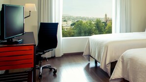 Quarto de hotel com cama, escrivaninha com computador, cadeira e janela com vista para a cidade.