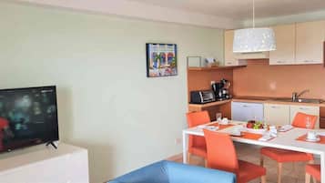 Espaço compacto para viver, com televisão, mesa de jantar, cadeiras, cozinha e um pequeno sofá.