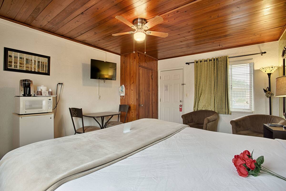 Quarto de hotel com teto de madeira, uma cama com edredom branco, escrivaninha com micro-ondas, televisão na parede e janela com cortinas.