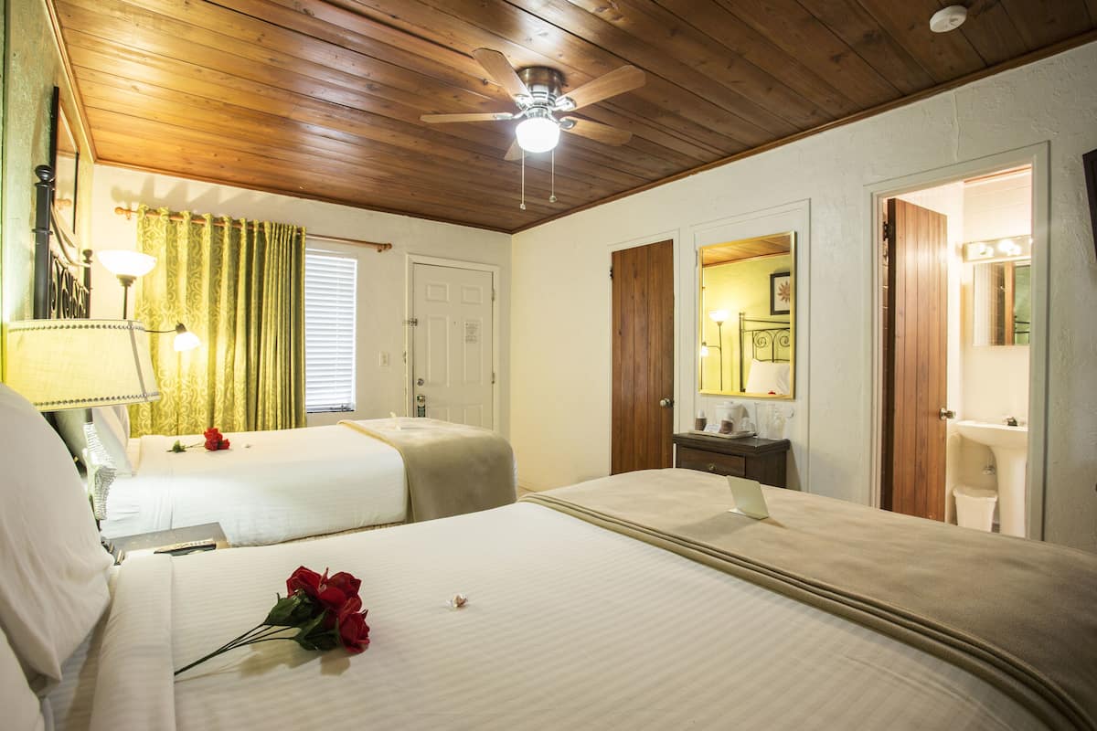 Quarto de hotel com duas camas, um ventilador de teto, uma janela com cortinas, uma mesinha de cabeceira com abajur e um banheiro visível através de uma porta aberta.
