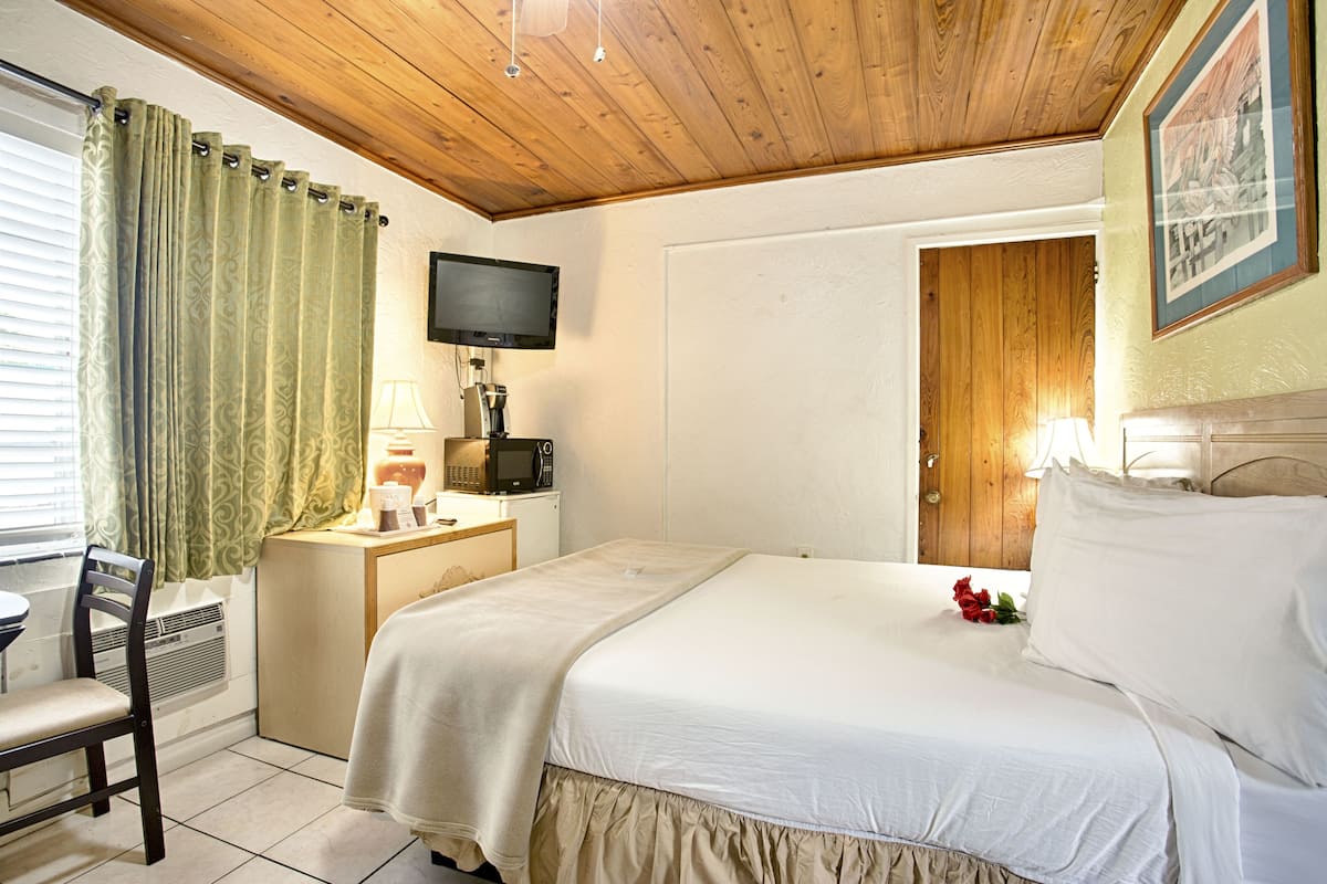 Quarto de hotel com teto de madeira, cama com lençóis brancos, escrivaninha com abajur, televisão e janela com cortinas.