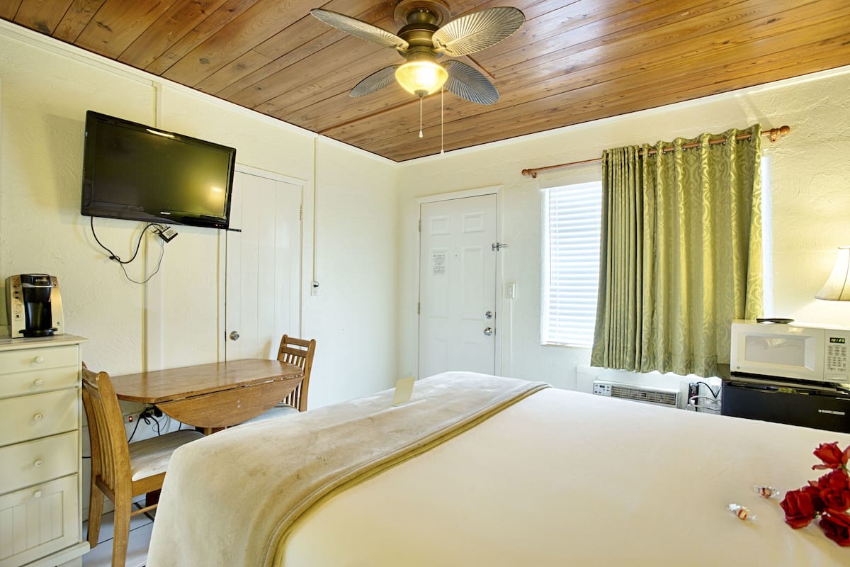 Quarto com cama, escrivaninha, cadeira, televisão, ventoinha no teto, janela com cortinas e micro-ondas.