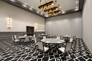Uma sala de conferência com mesas redondas e cadeiras, um tapete estampado e um lustre no teto.