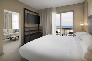 Quarto de hotel com uma cama grande, televisão de tela plana fixada na parede, varanda com vista para o oceano e área de estar com sofá e poltronas.