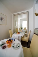 Uma sala de jantar com duas mesas, cada uma com toalhas brancas, pratos e tigelas de fruta. Um espelho grande reflete o espaço, e há uma moldura com um quadro na parede.
