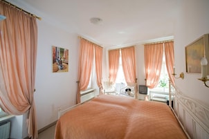 Quarto com uma cama grande, duas janelas com cortinas, uma pequena mesa com cadeira, televisão e um quadro na parede.