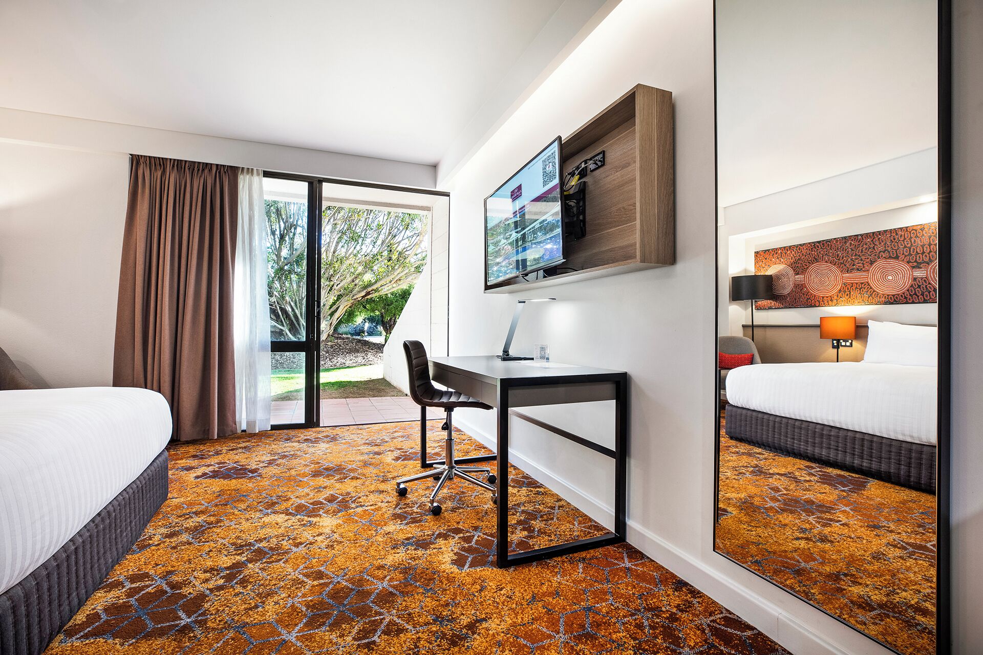 Quarto de hotel com cama, escrivaninha com cadeira, televisão na parede e um espelho grande refletindo o ambiente.