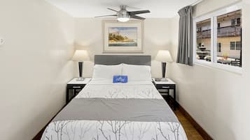 Quarto de hotel com cama, duas mesas de cabeceira, ventilador de teto, janela com vista para uma varanda e um quadro na parede.