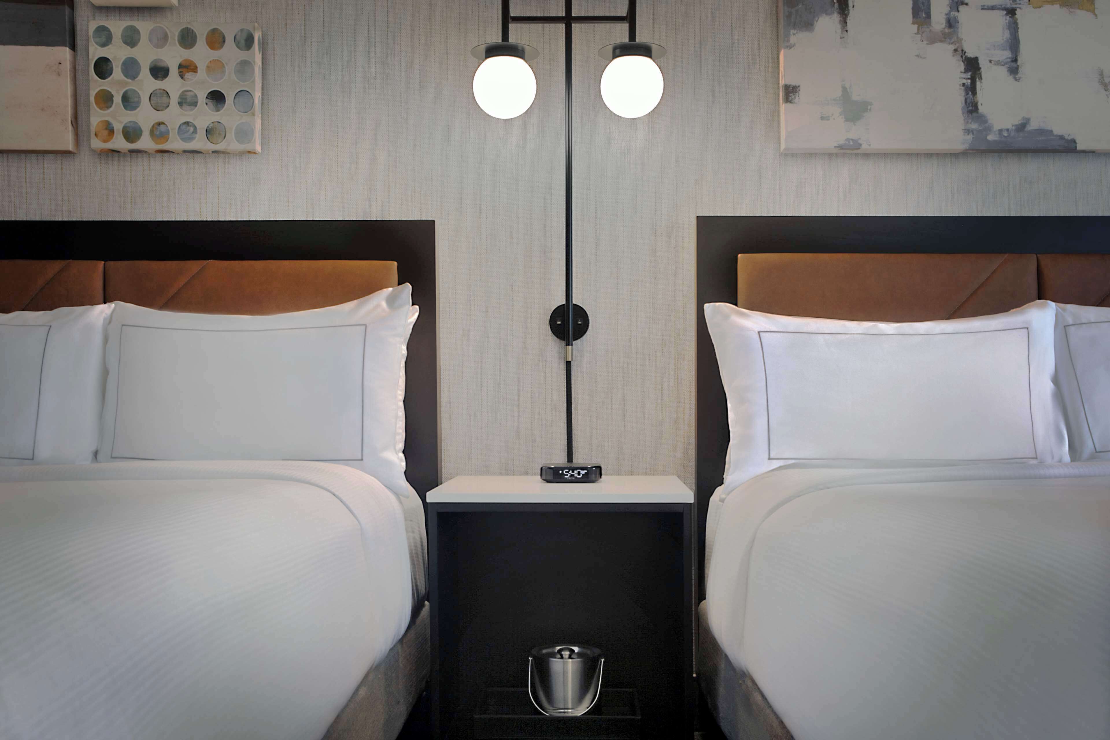 Quarto de hotel com duas camas, uma mesa de cabeceira com relógio e uma luminária fixada na parede.