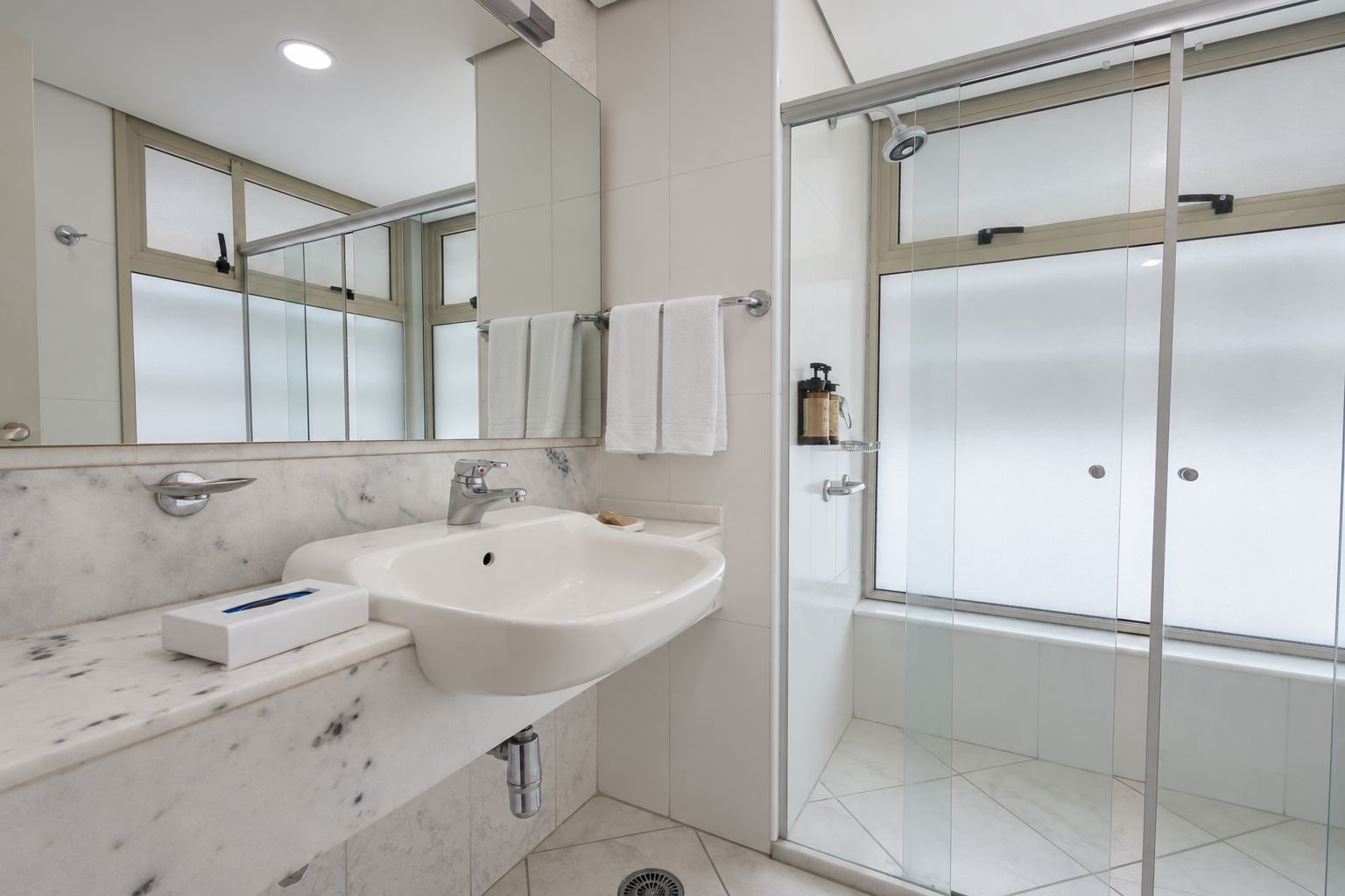 Casa de banho moderna com bancada de mármore, lavatório branco, cabine de duche envidraçada e espelho grande.
