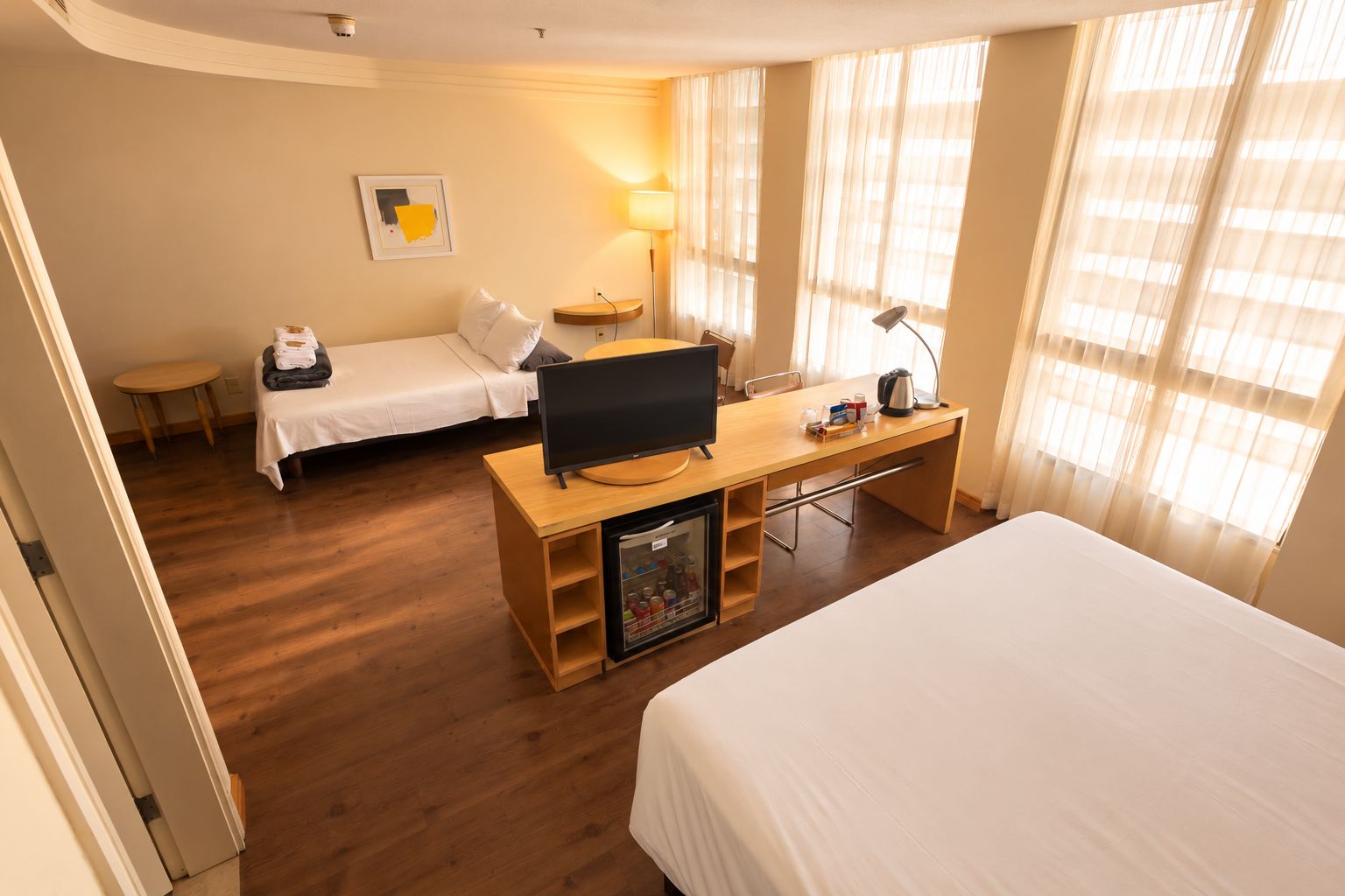 Quarto de hotel com cama, escrivaninha com computador, mini-frigorífico e janela com cortinas.