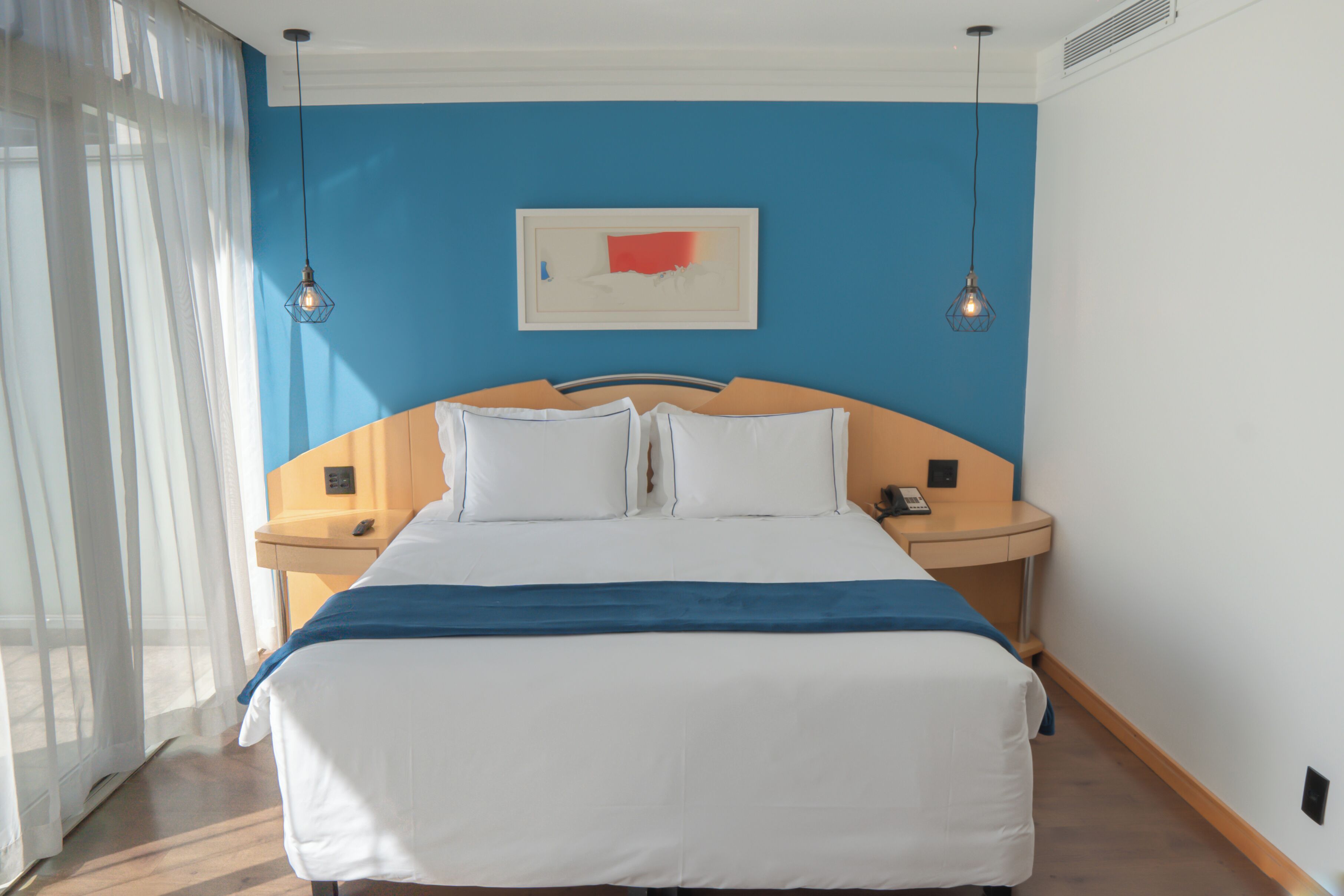 Quarto de hotel com cama, mesas de cabeceira, um criado-mudo, um quadro na parede e cortinas finas na janela.