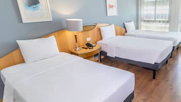 Quarto de hotel com três camas de solteiro, cabeceira de madeira, mesa de cabeceira com abajur, telefone e quadros nas paredes.