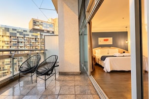 Uma varanda com duas cadeiras, uma cama com roupa de cama branca e vista para a cidade.