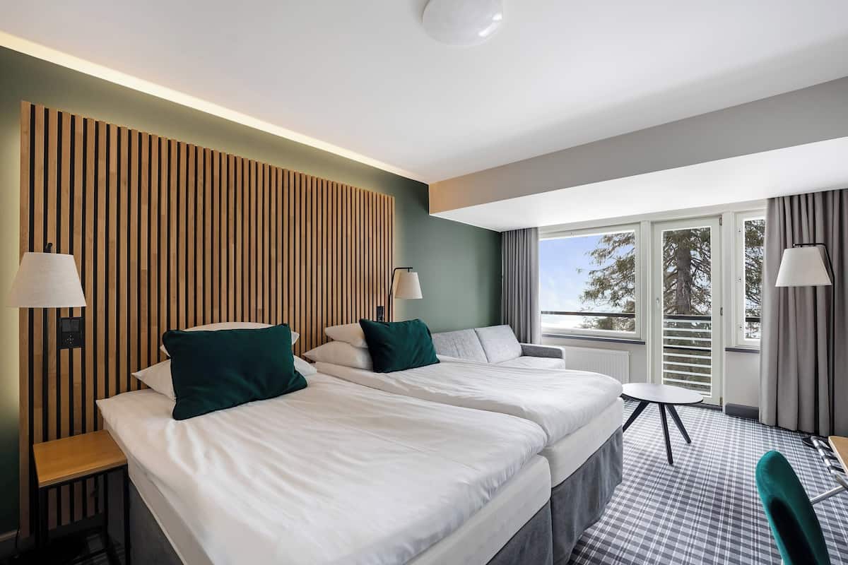 Quarto de hotel com duas camas, uma mesa pequena e uma cadeira. Há uma janela com vista para árvores e uma varanda.