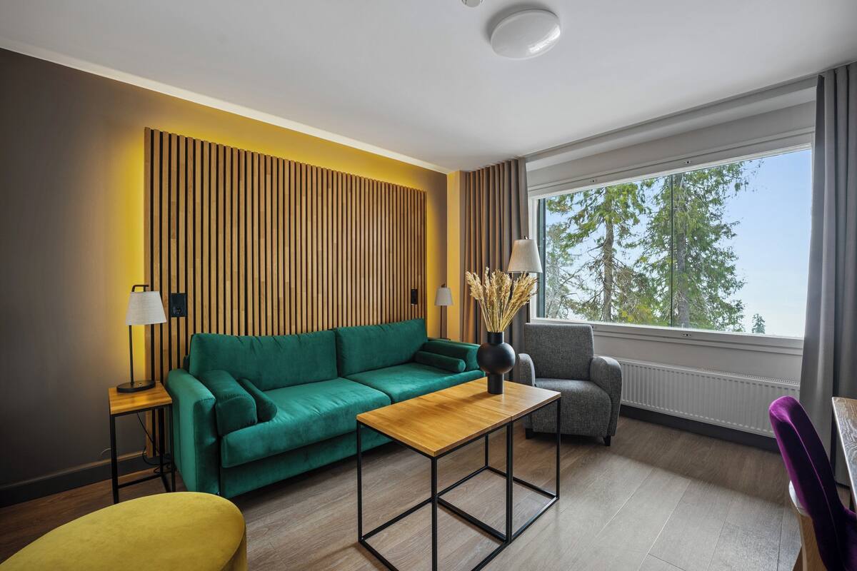 Uma sala de estar moderna com um sofá verde, uma mesa de centro de madeira e uma poltrona cinza. Uma luminária e um vaso com plantas secas estão na mesa lateral.