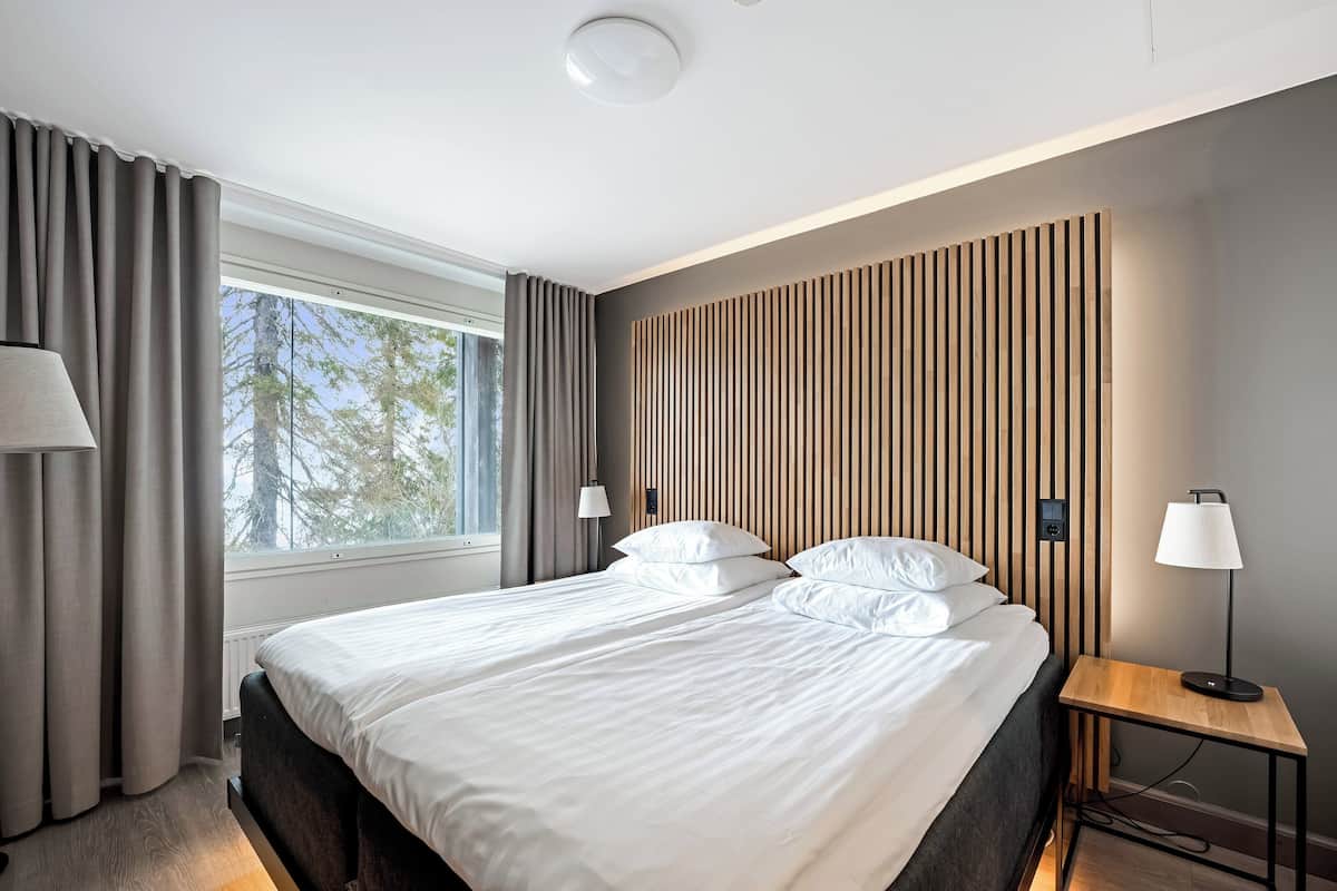 Quarto de hotel moderno com uma cama grande, duas luminárias de cabeceira, uma mesa de apoio e uma janela com cortinas.