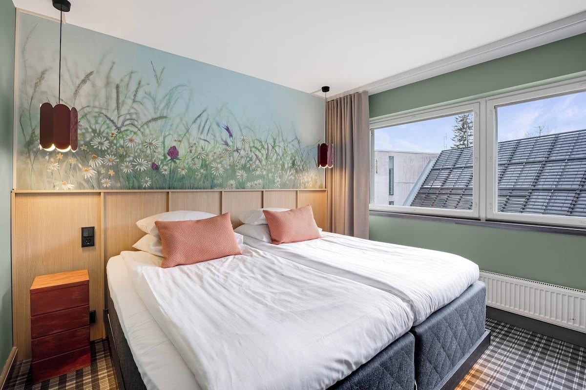 Quarto moderno com uma cama grande, um mural na parede, uma janela com vista para edifícios e uma mesa de cabeceira.