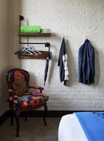 Um quarto com uma cadeira de madeira, uma cama, uma parede de tijolos, um cabideiro e uma estante com uma toalha verde e livros.