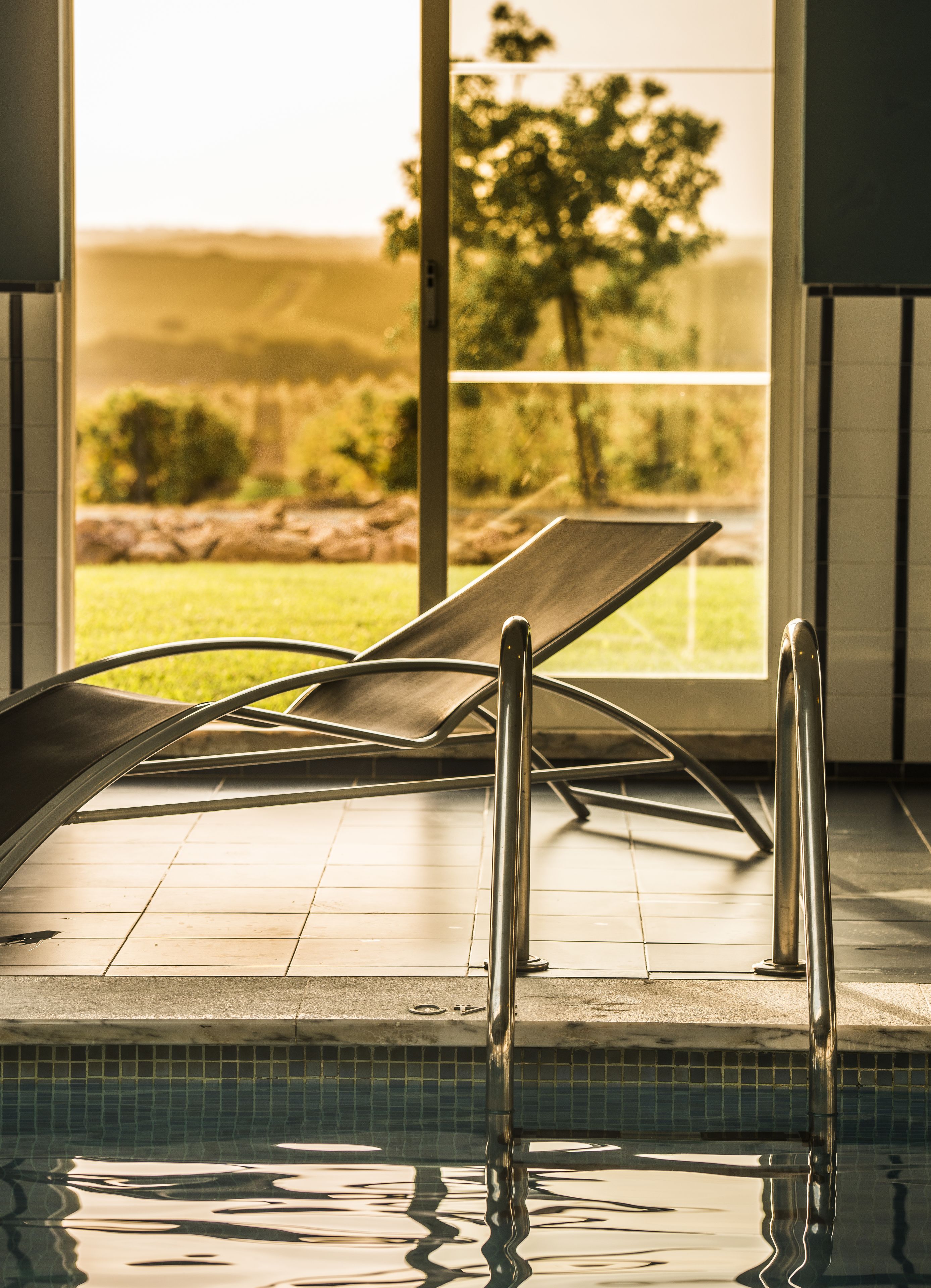 Uma área à beira da piscina com espreguiçadeira e grades de metal, chão de azulejos e vista para a paisagem através de grandes janelas.