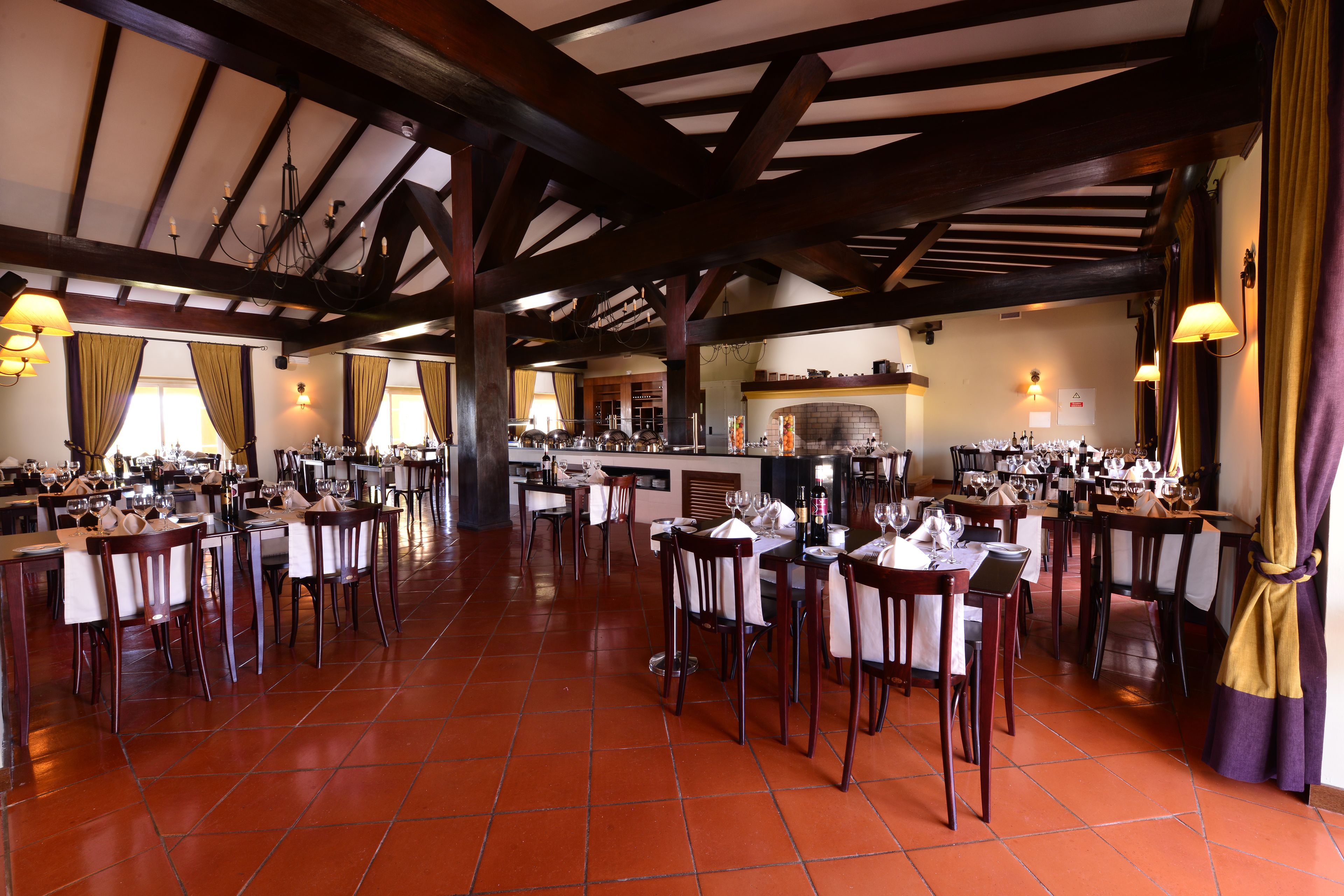 Uma sala de jantar ampla com várias mesas e cadeiras, vigas de madeira no teto e piso de tijolo.