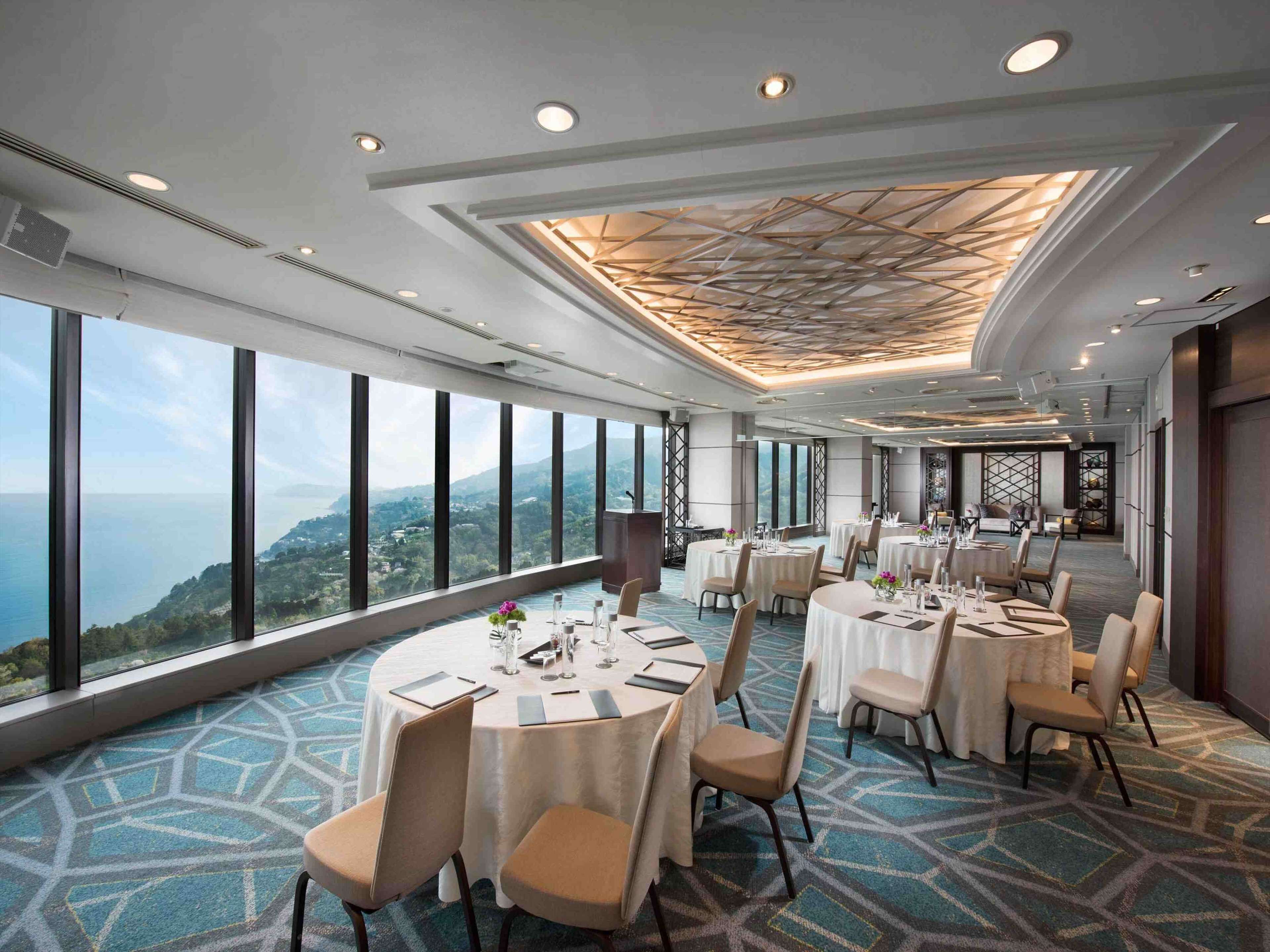 Uma sala de jantar ampla, com pé direito alto, mesas redondas, grandes janelas com vista panorâmica e um tapete estampado.