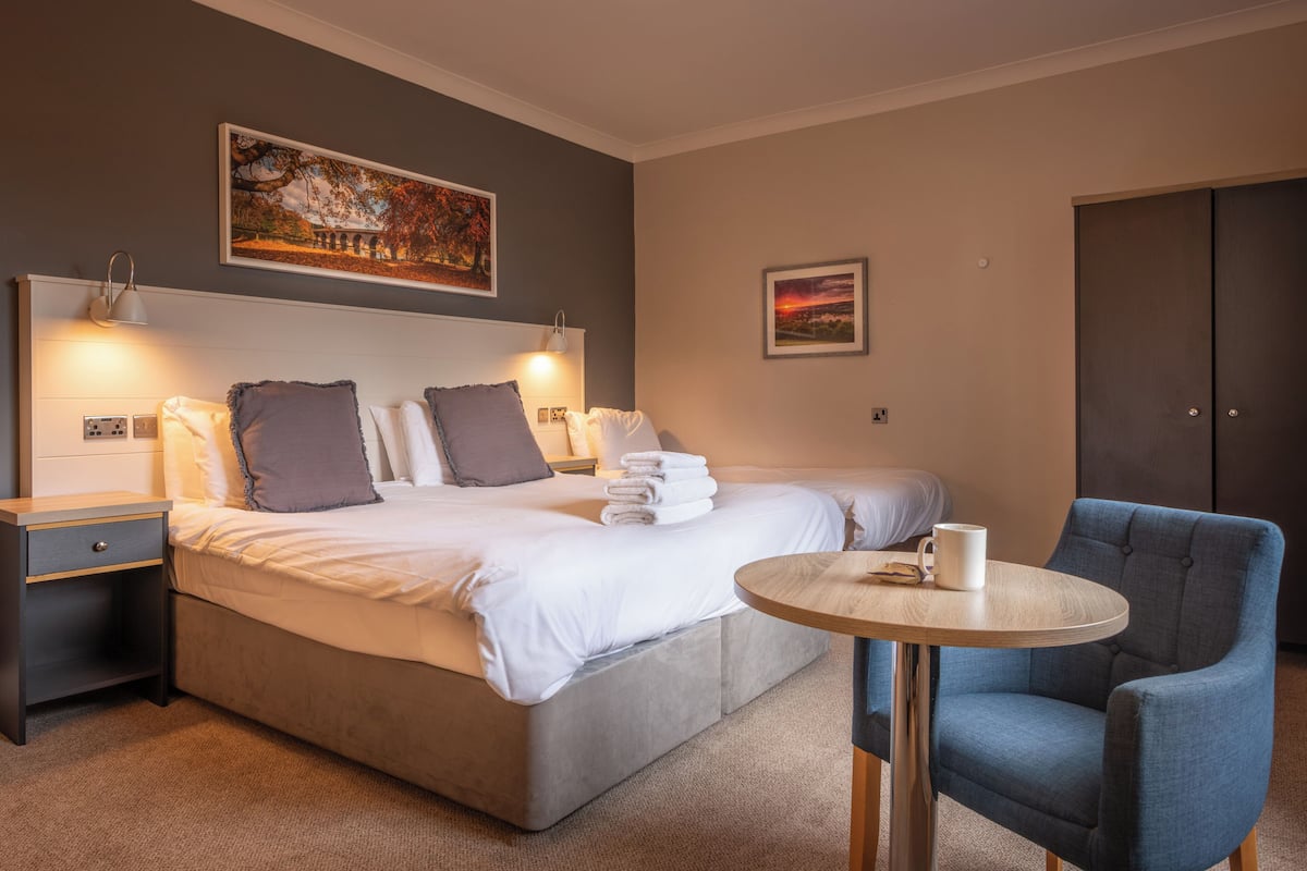 Quarto de hotel com cama de casal, mesas de cabeceira, uma mesinha redonda com cadeira e duas luminárias de parede.
