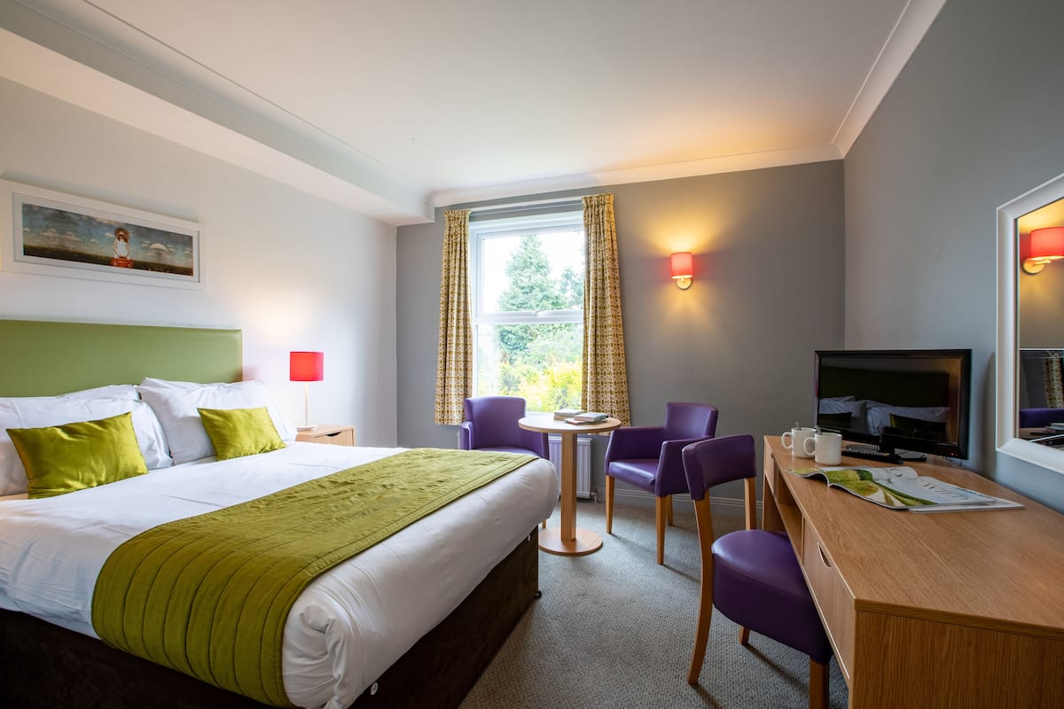 Quarto de hotel com cama, escrivaninha, cadeira, televisão e janela com cortinas.
