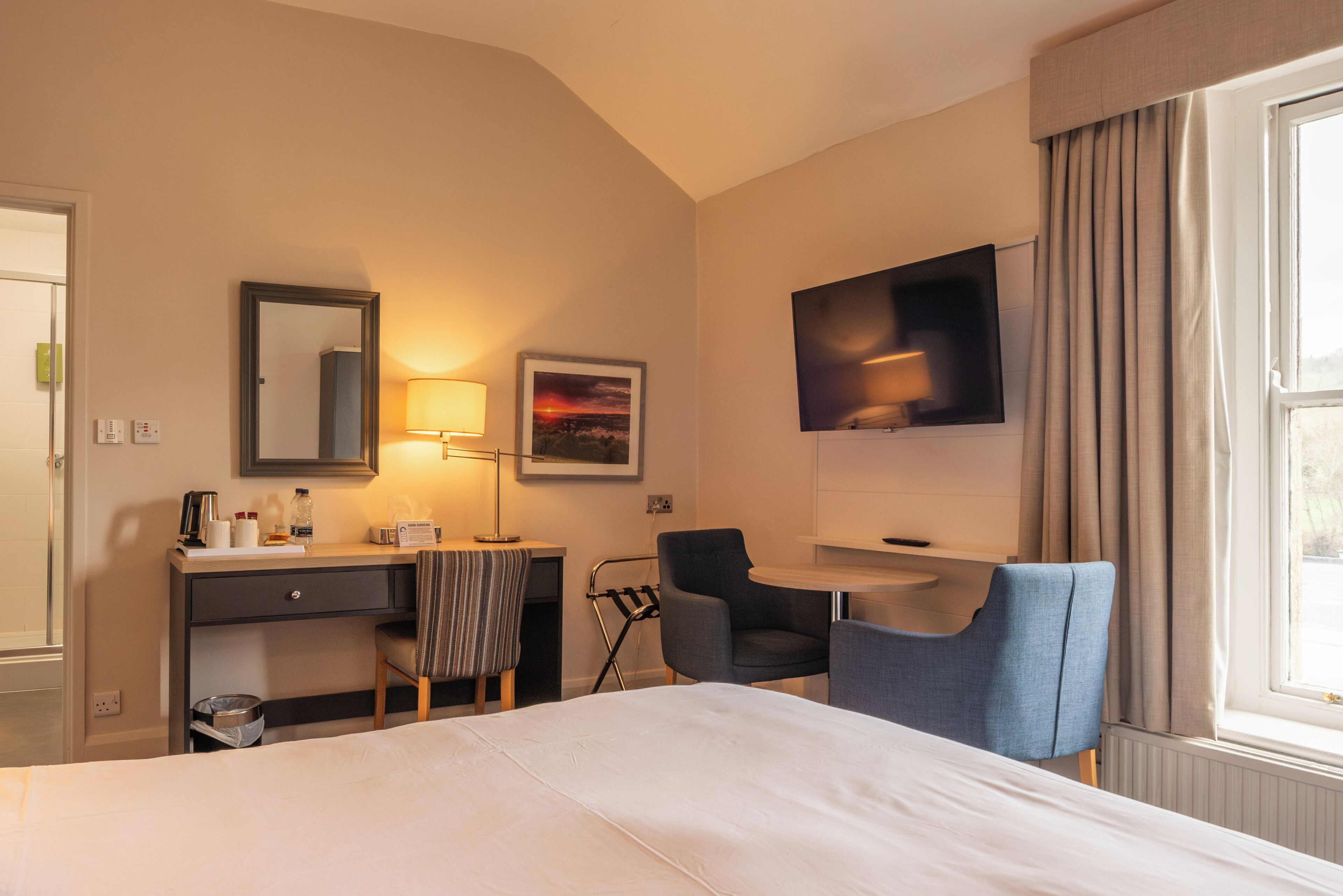 Quarto de hotel com cama, escrivaninha, cadeira, televisão e janela com cortinas.