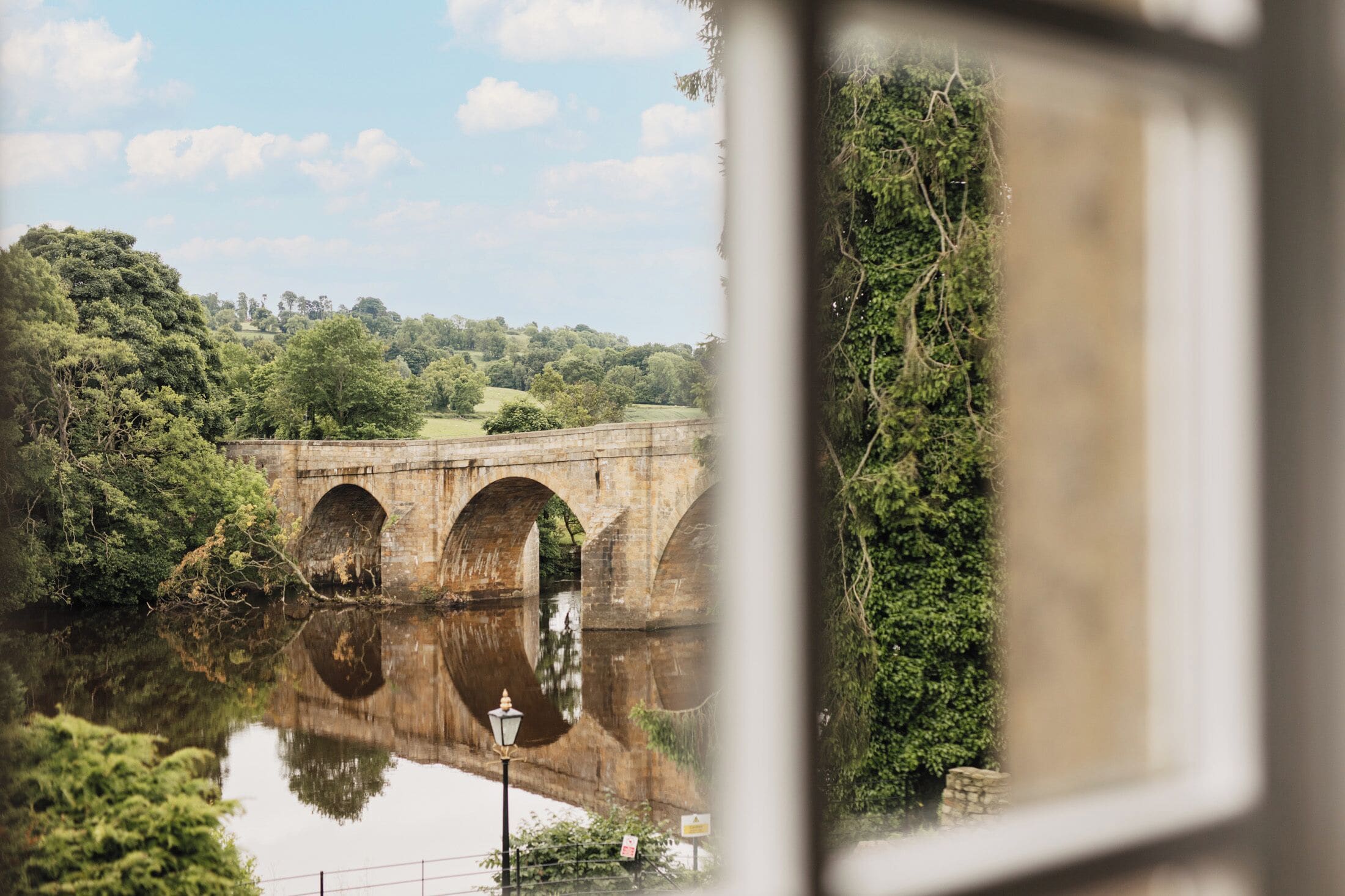 Uma ponte de pedra sobre um rio calmo, rodeada por vegetação exuberante.