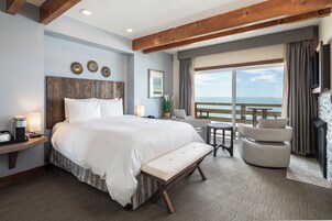 Quarto de hotel com uma cama grande, um banco, uma escrivaninha com abajur, uma cadeira e vista para o mar.