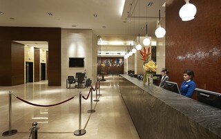 Uma área de recepção de hotel com um balcão de mármore, uma televisão na parede e uma corda de veludo delimitando a área.