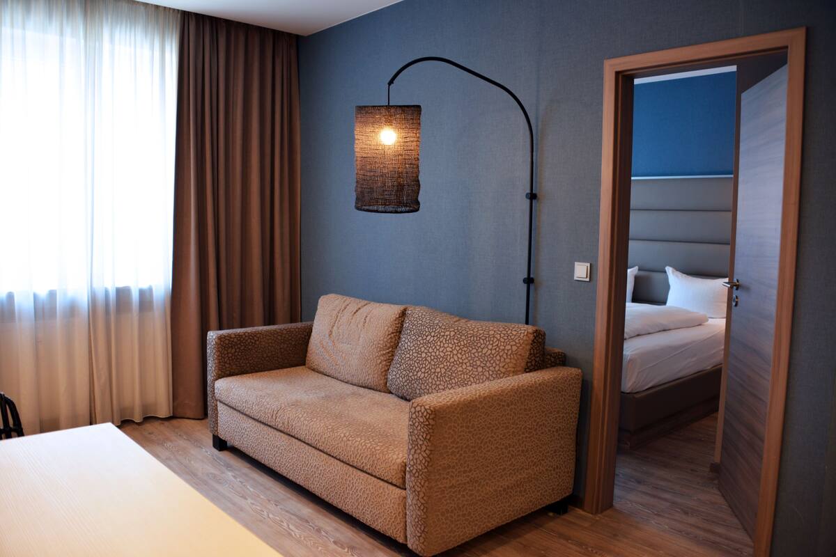 Quarto de hotel com sofá, mesa de cabeceira, abajur e cama.