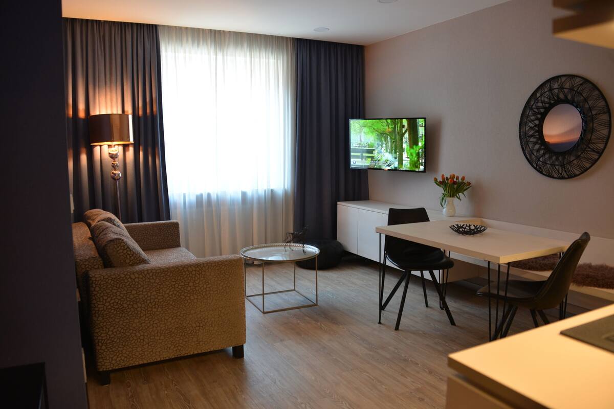 Uma sala de estar moderna com sofá, mesa de centro, mesa de jantar com cadeiras, TV fixada na parede e janela com cortinas.