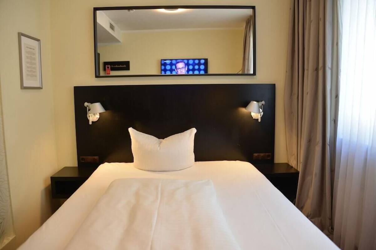 Quarto de hotel com uma cama grande, cabeceira com duas luminárias fixadas na parede, televisão de tela plana e espelho sobre a cama.
