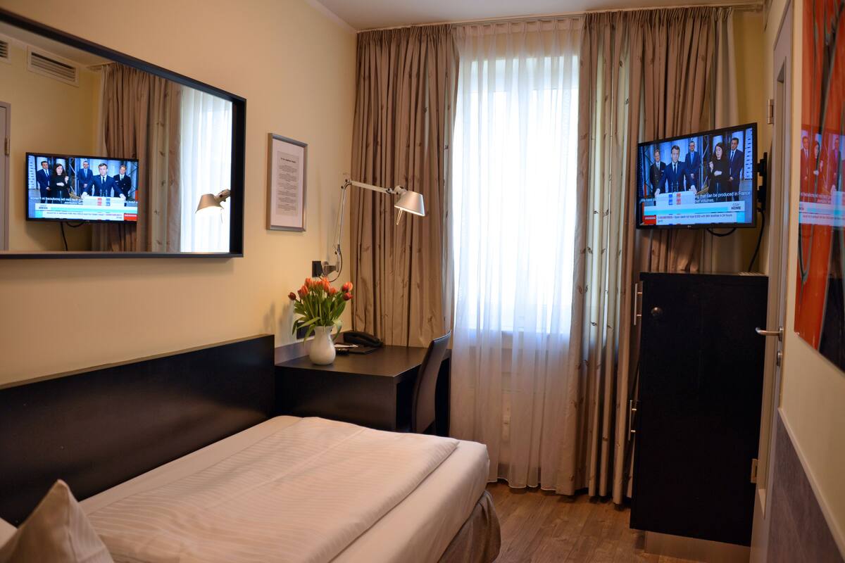 Quarto de hotel com uma cama, uma escrivaninha com luminária, uma televisão e uma janela com cortinas.