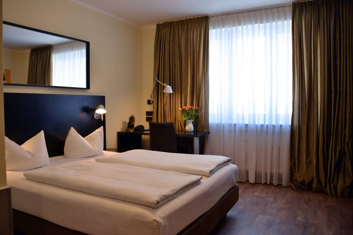 Quarto de hotel com duas camas, uma escrivaninha, uma cadeira, uma luminária e uma janela grande com cortinas.