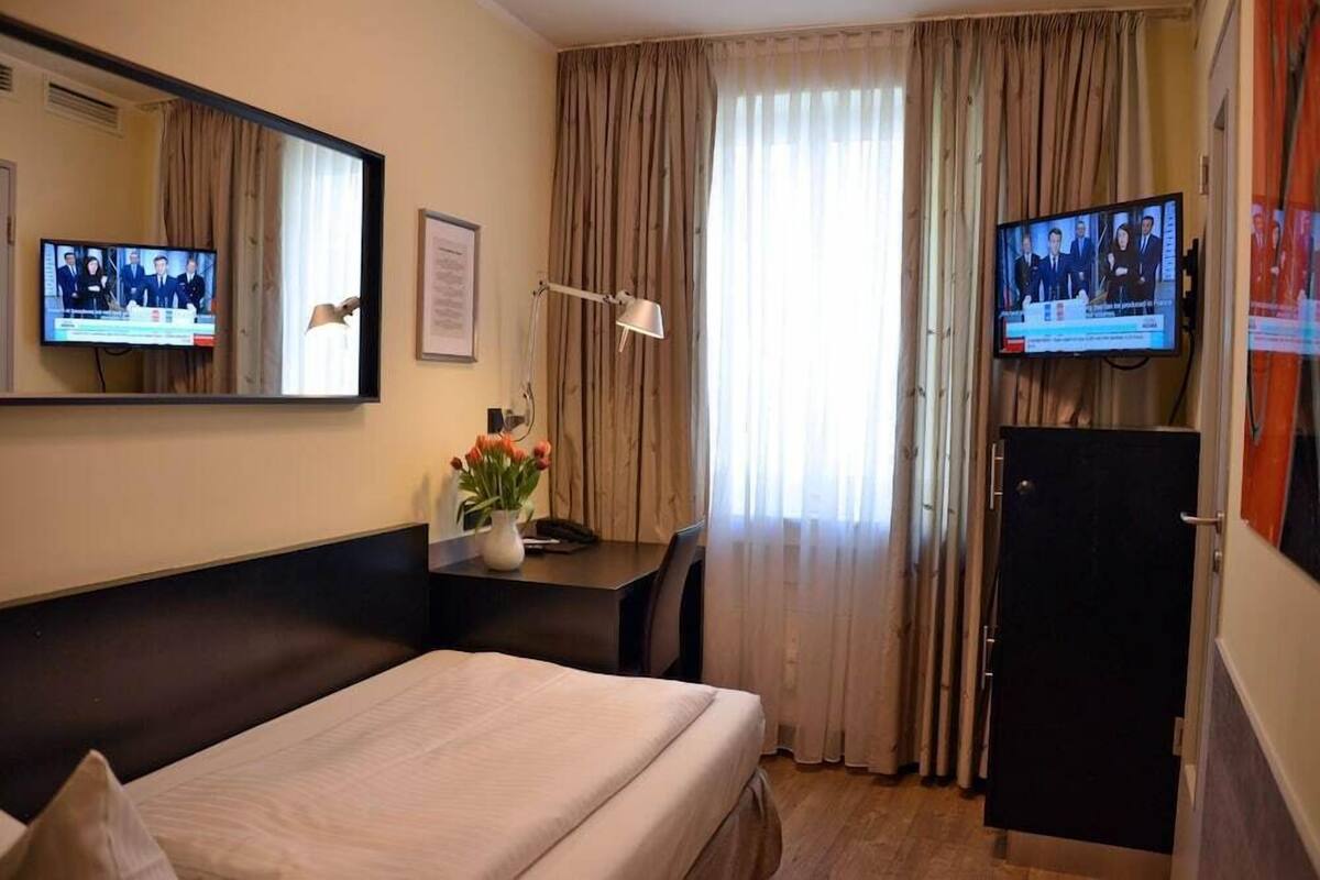 Quarto de hotel com uma cama, uma mesa com luminária, uma televisão e uma janela com cortinas.