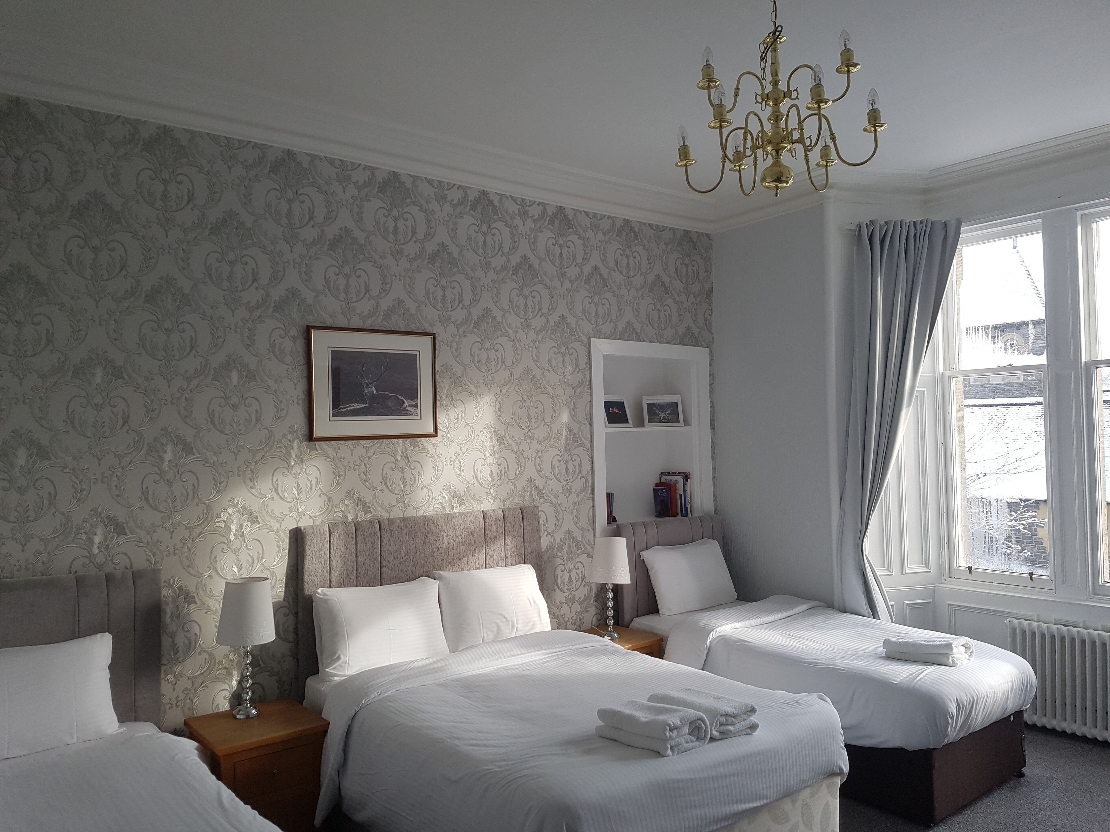 Quarto de hotel com duas camas, um candelabro, uma janela com cortinas e um papel de parede estampado.