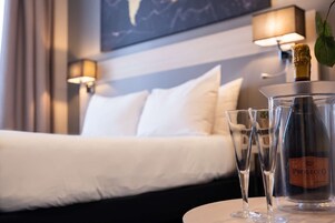 Quarto de hotel com uma cama, duas luminárias de cabeceira, uma garrafa de Prosecco e dois copos sobre uma mesa.
