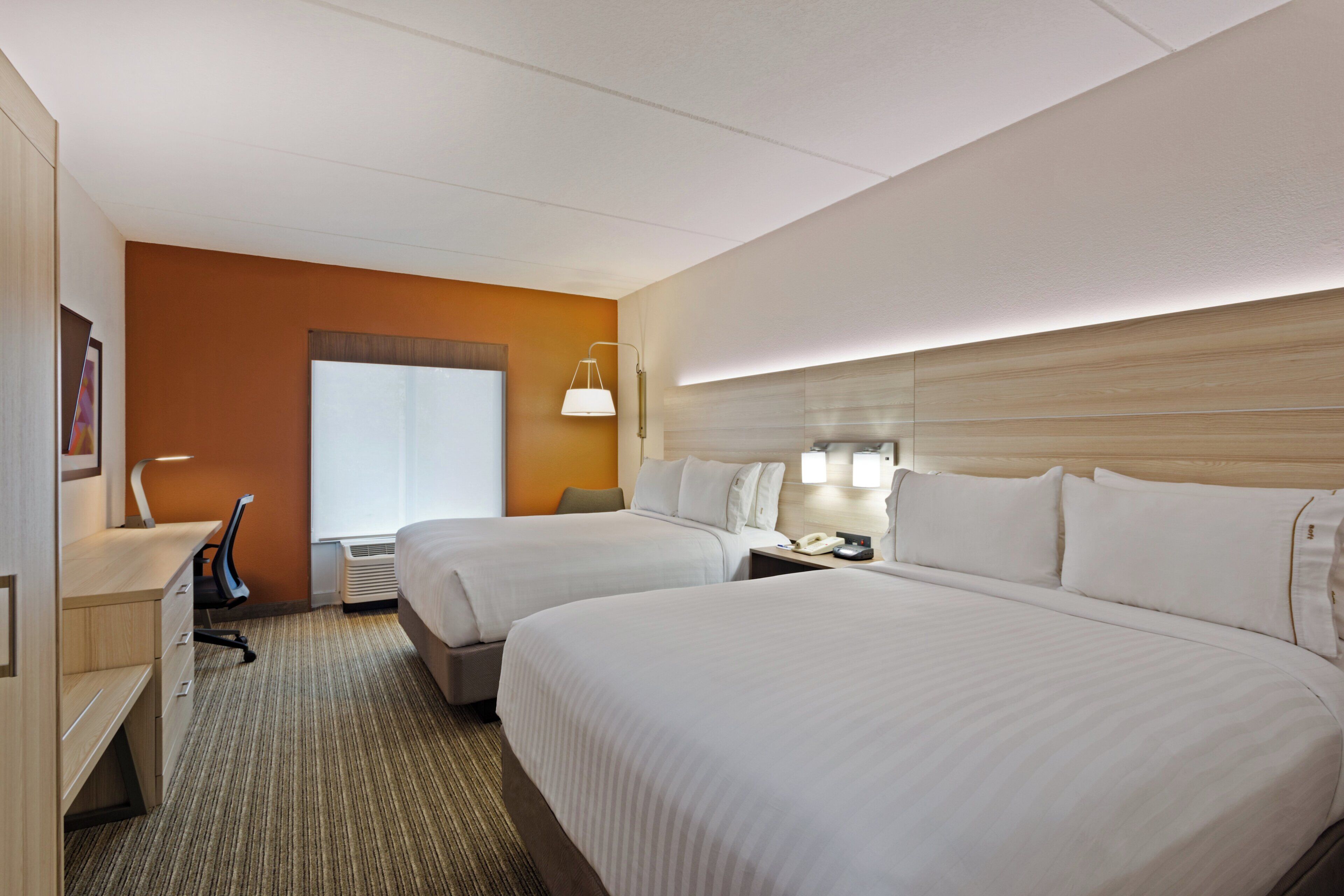Quarto de hotel com duas camas, uma escrivaninha, uma cadeira, um abajur e uma janela.