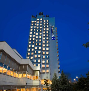 Um hotel Radisson imponente, com vários andares e varandas, exibindo uma placa chamativa com o nome 'Radisson' e o logotipo 'BLU'.