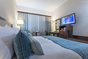 Quarto de hotel com cama, escrivaninha, cadeira, televisão e janela com cortinas.
