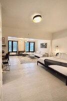 Uma sala de estar espaçosa com uma cama grande, um sofá, uma mesa de jantar e uma mesa de centro. Há janelas amplas com cortinas, um lustre e um quadro na parede.