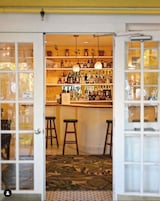 Uma área de bar com prateleiras de garrafas de bebidas, dois bancos e luminárias pendentes.
