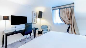 Quarto de hotel com uma cama, uma escrivaninha com cadeira, TV de tela plana, abajur e janela com cortinas.