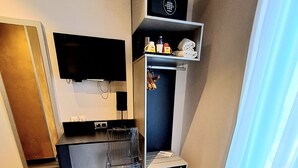 Quarto de hotel com cama, televisão na parede, pequena escrivaninha com cadeira, prateleira com amenities de higiene pessoal e janela com cortinas.